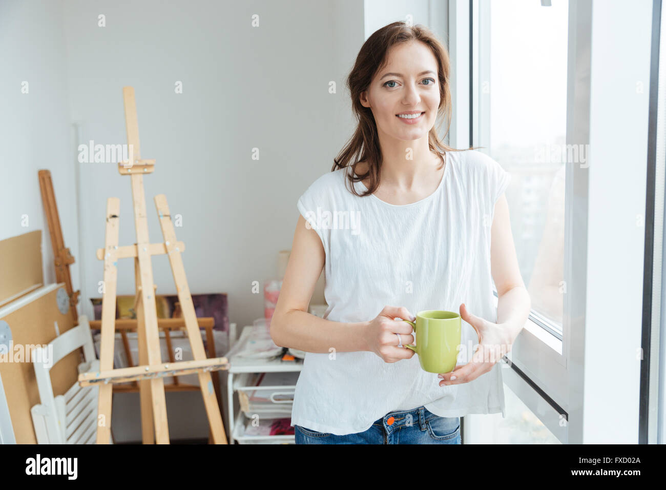 Cheerful beautiful woman painter drinking tea near the window in art ...