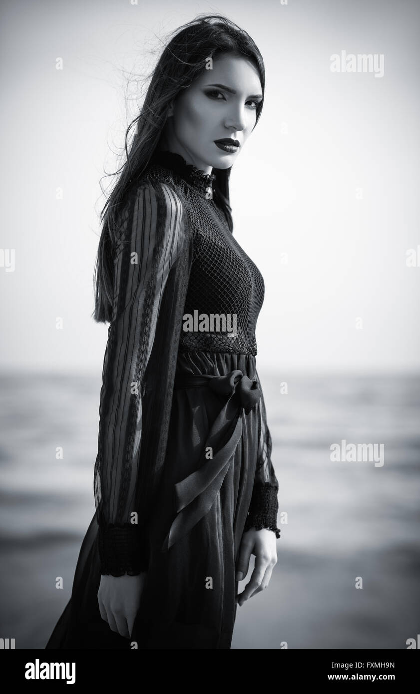 Beautiful sad gothic woman in black dress standing on the sea beach ...