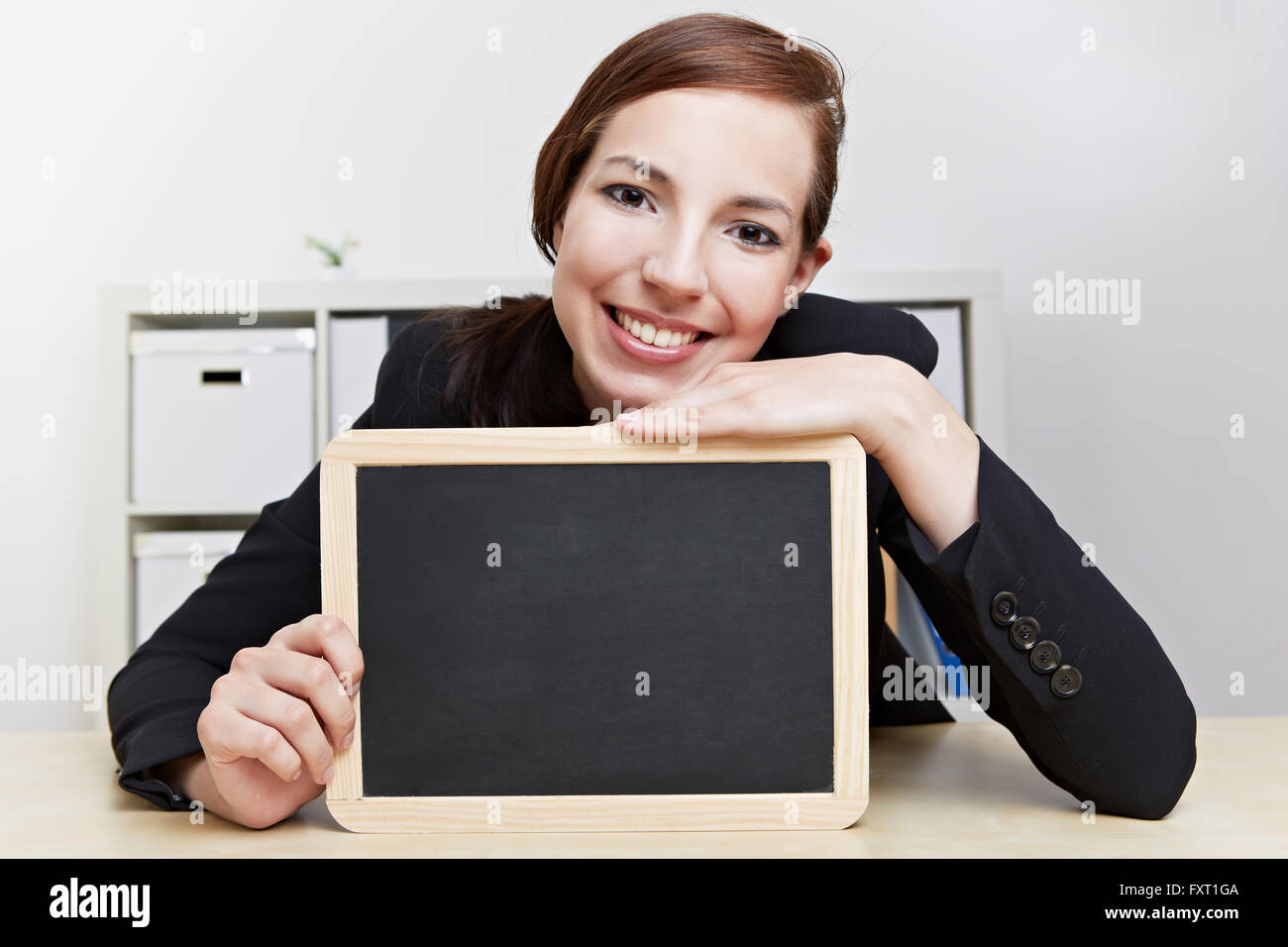 Smiling Female Teacher Sitting With Empty Black Chalkboard At Her Desk