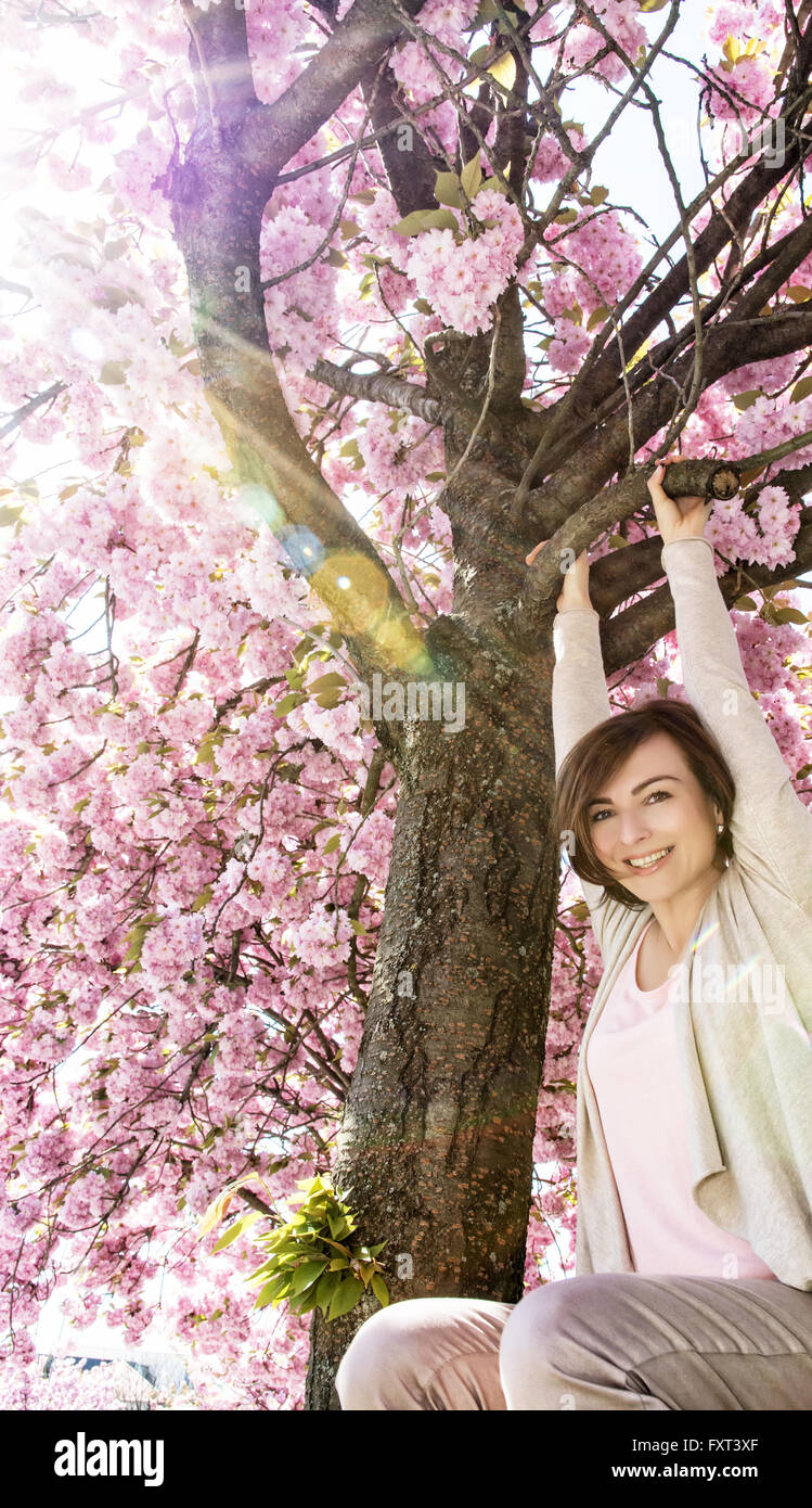 Happy young woman is hanging on the sakura tree with reflected sun rays ...