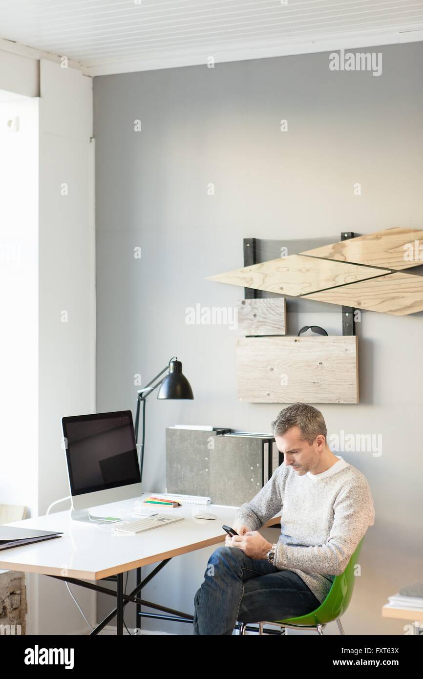 Mature man sitting at desk in office looking down using smartphone ...