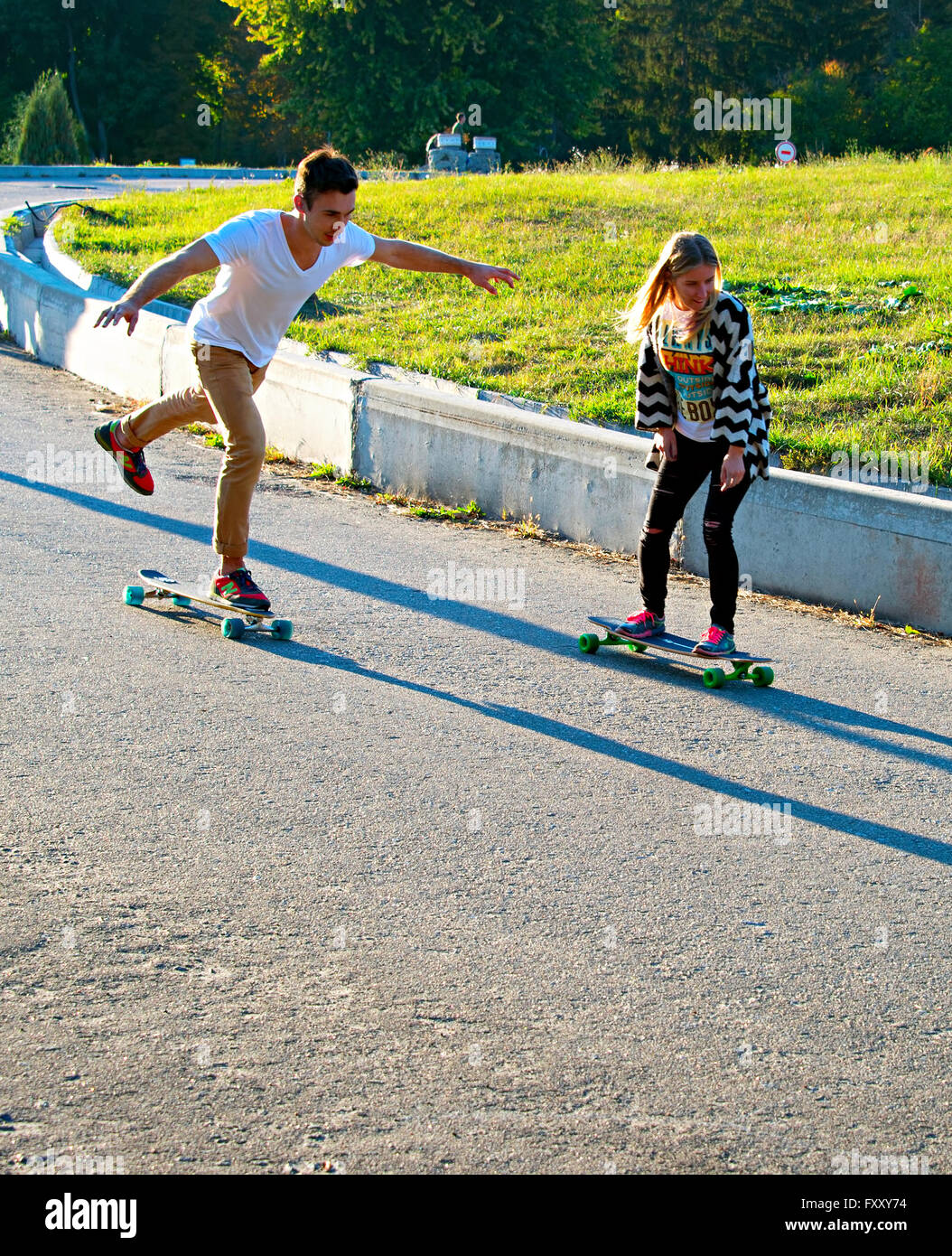 Young couple riding the longboards down the hill at sunset in a park ...