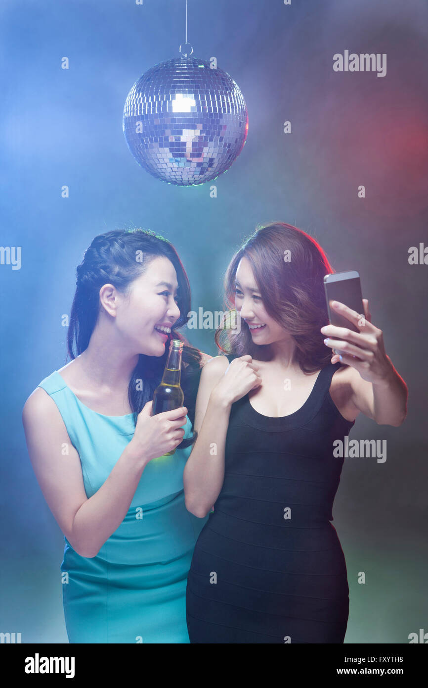 Two young smiling women with beer bottle and smart phone face ro face ...