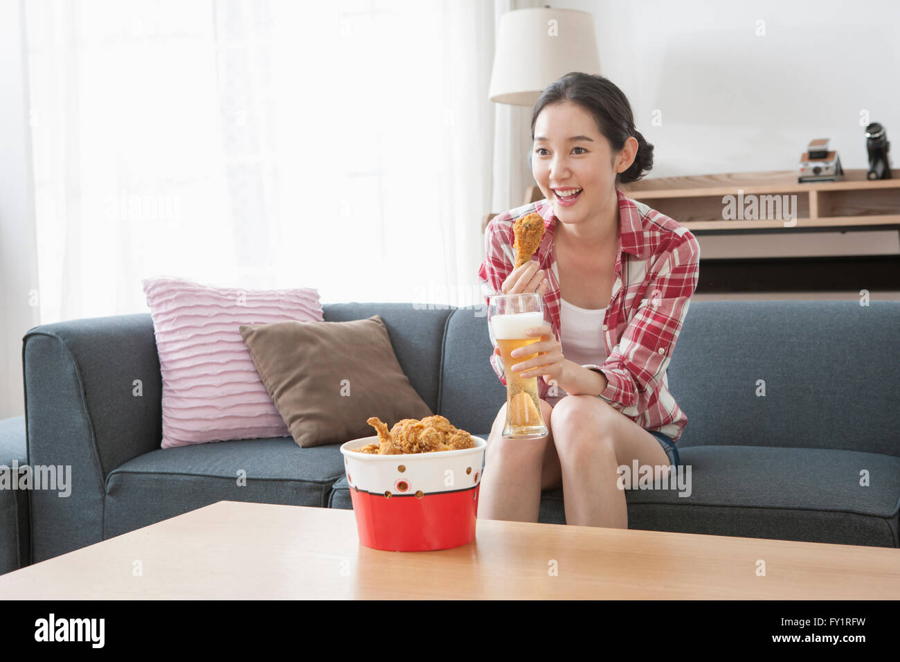 Young smiling woman sitting on sofa holding fried chicken and a glass ...