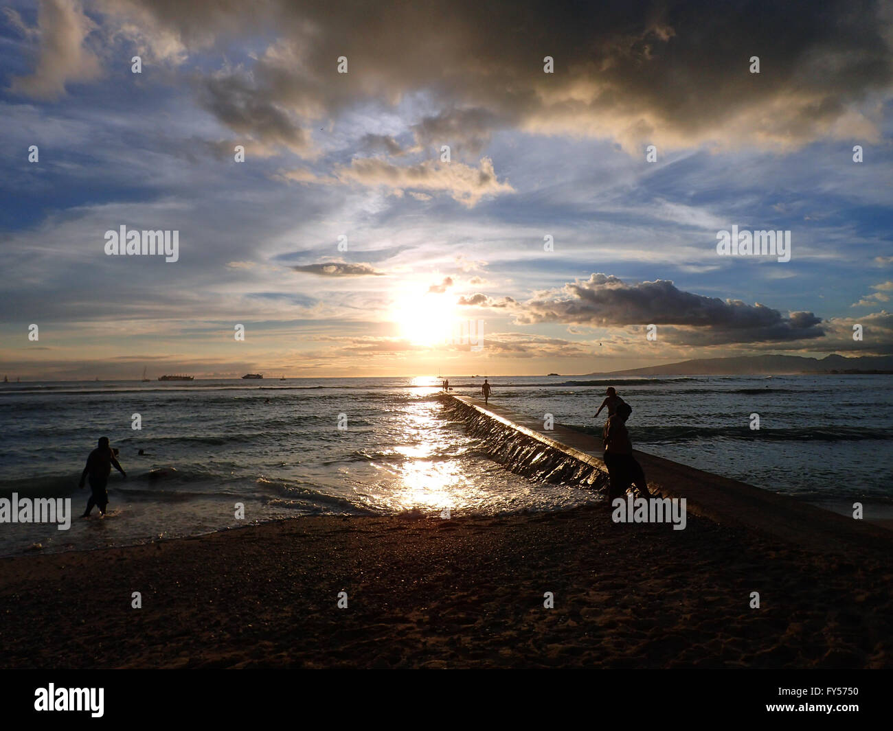 People walk along jetty path into the ocean as waves lap during Sunset ...