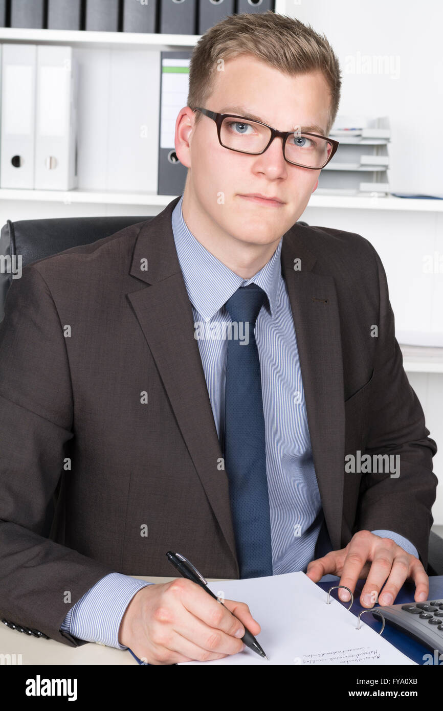 Young businessman is writing into a file while sitting at the desk in ...