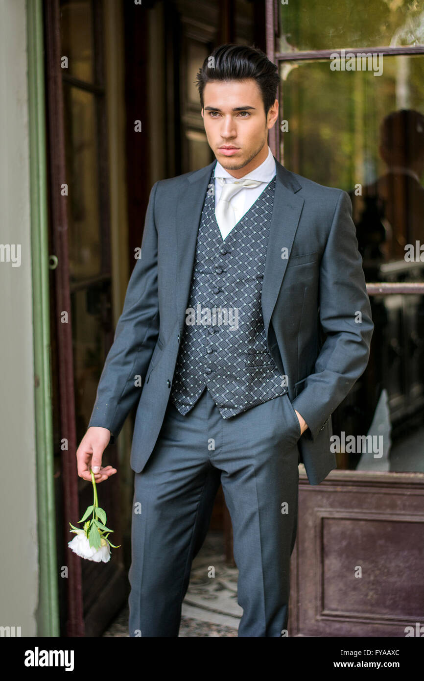 Portrait of elegant young bridgegroom in grey suit holding white flower ...
