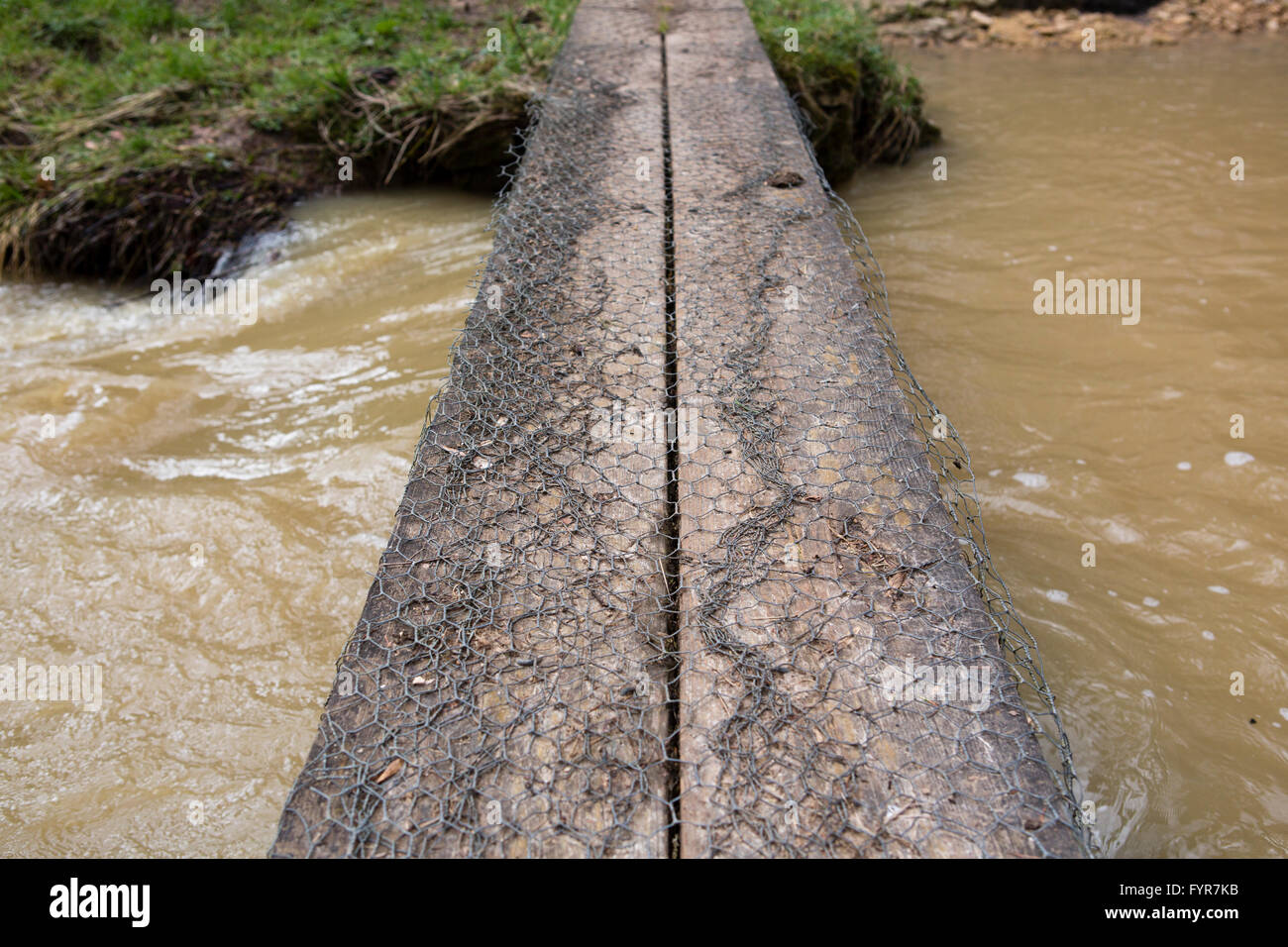 A swollen stream with muddy water flows under a narrow wooden bridge ...