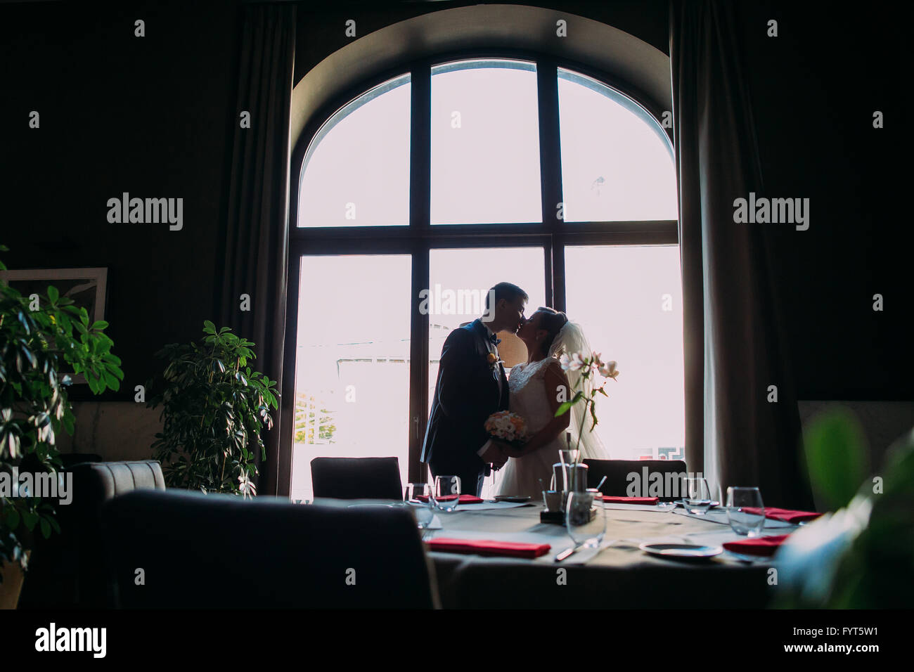 Beautiful young married couple standing by window kissing Stock Photo ...