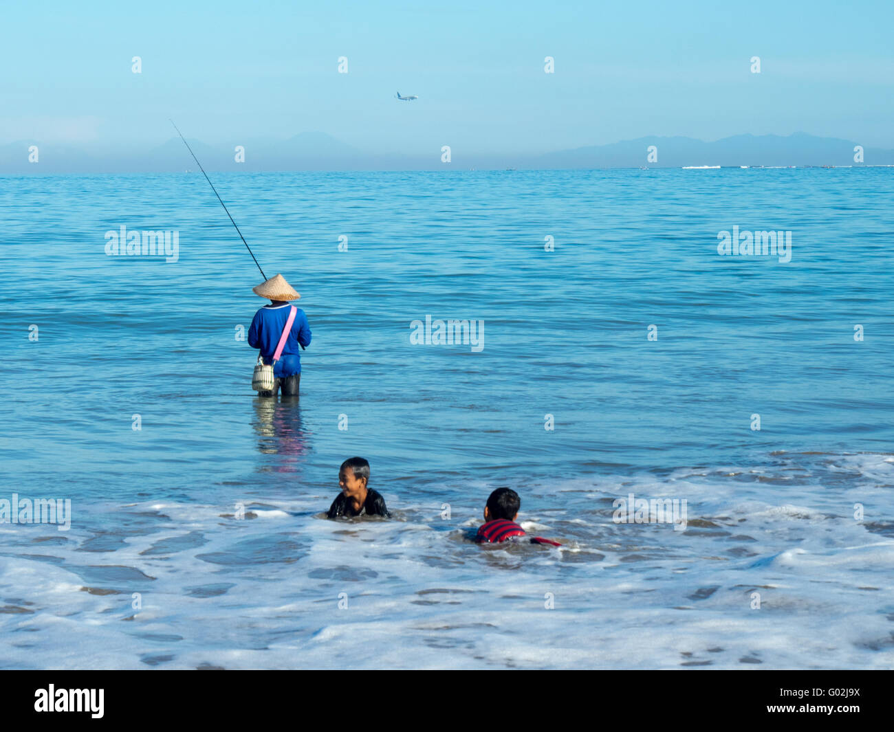 A man standing in the sea fishing while two young boys are playing ...
