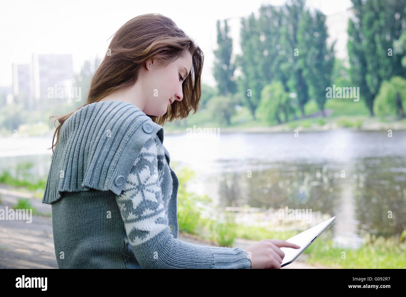Girl sitting outdoors using a touchscreen tablet along a riverbank ...