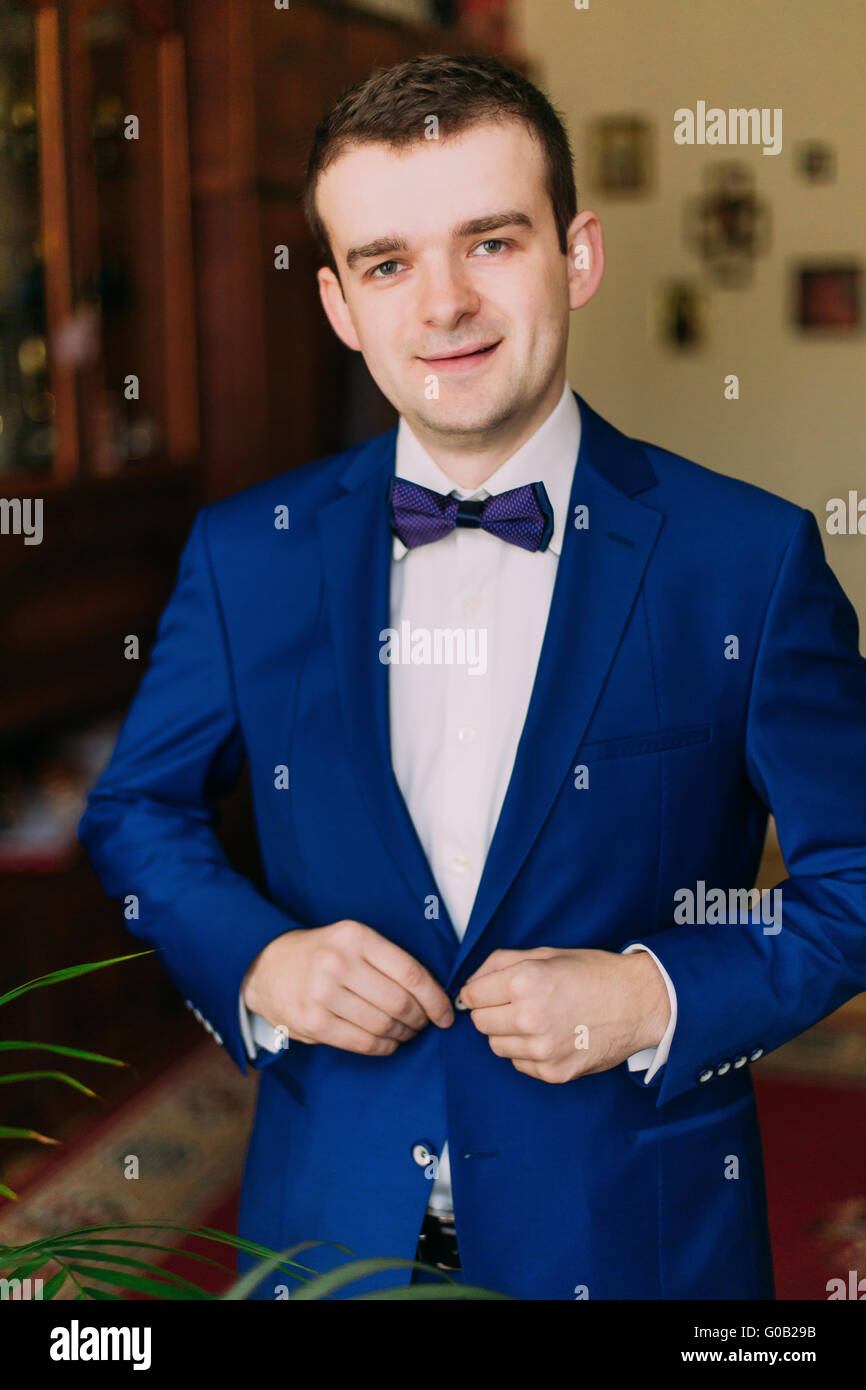 Adult solid man button his blue suit near window looking at the camera ...