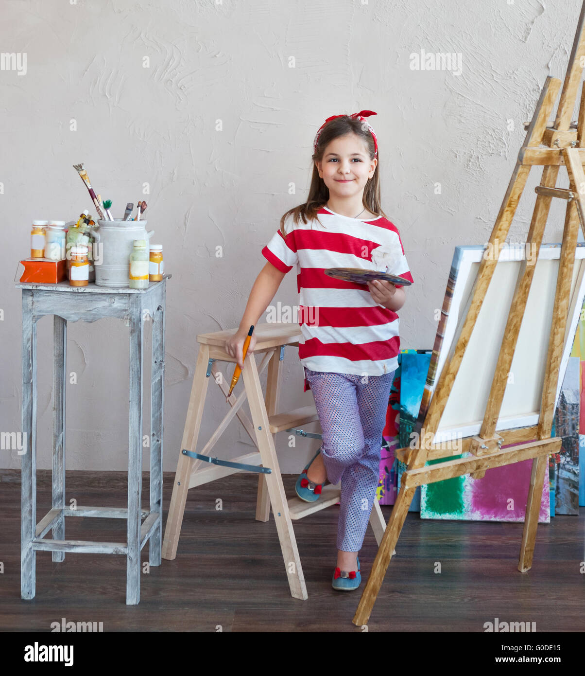 Little artist girl holding a paintbrush and looking over a canvas on an ...