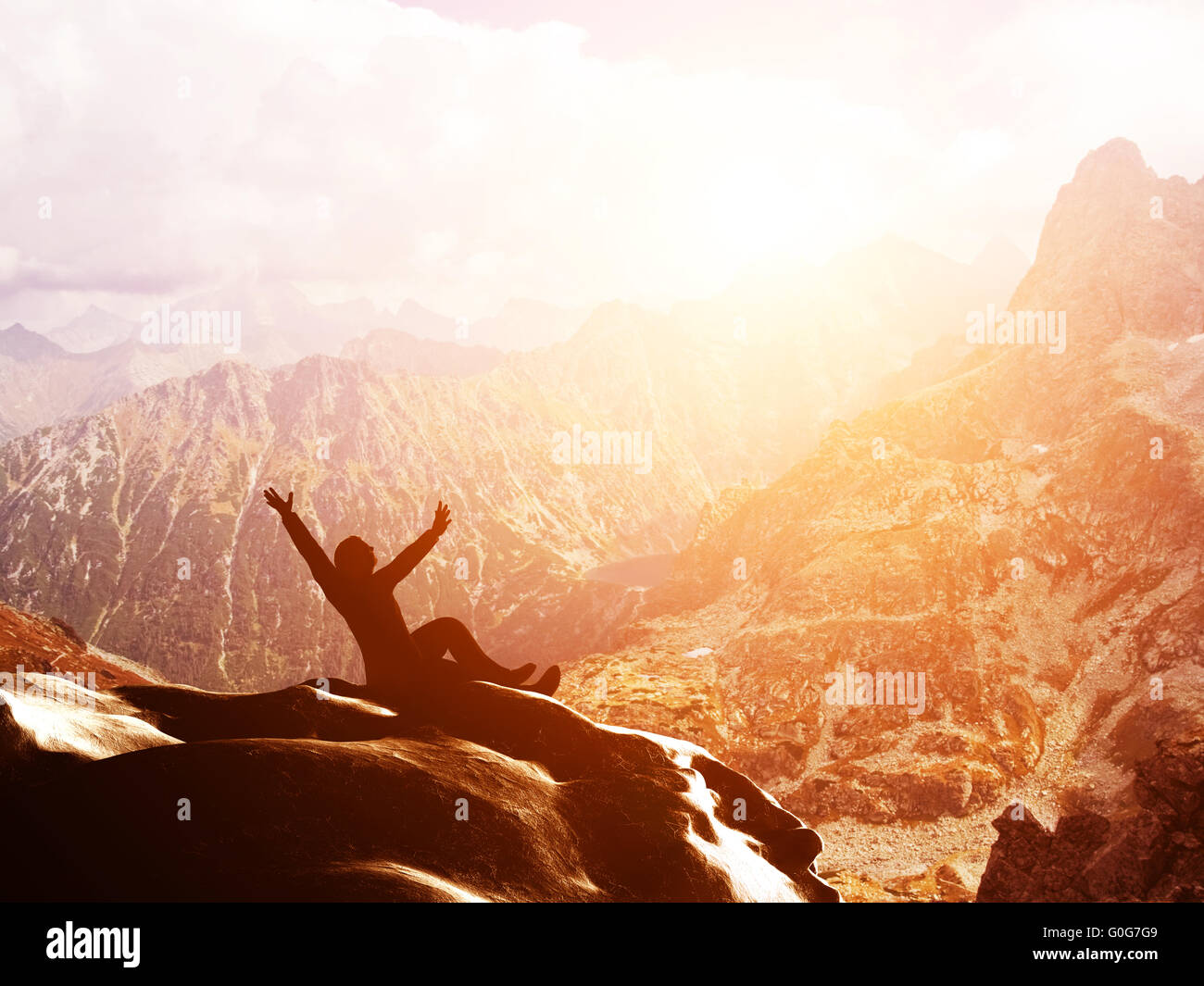 A happy man sitting on the peak of a mountain with hands raised ...
