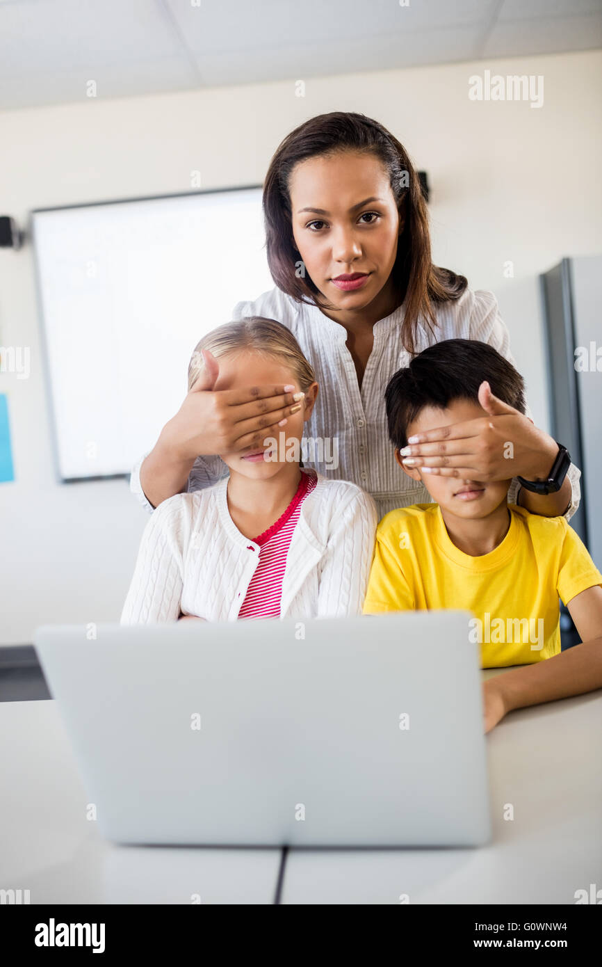 Teacher covering pupils eyes in front of computer and looking at camera ...