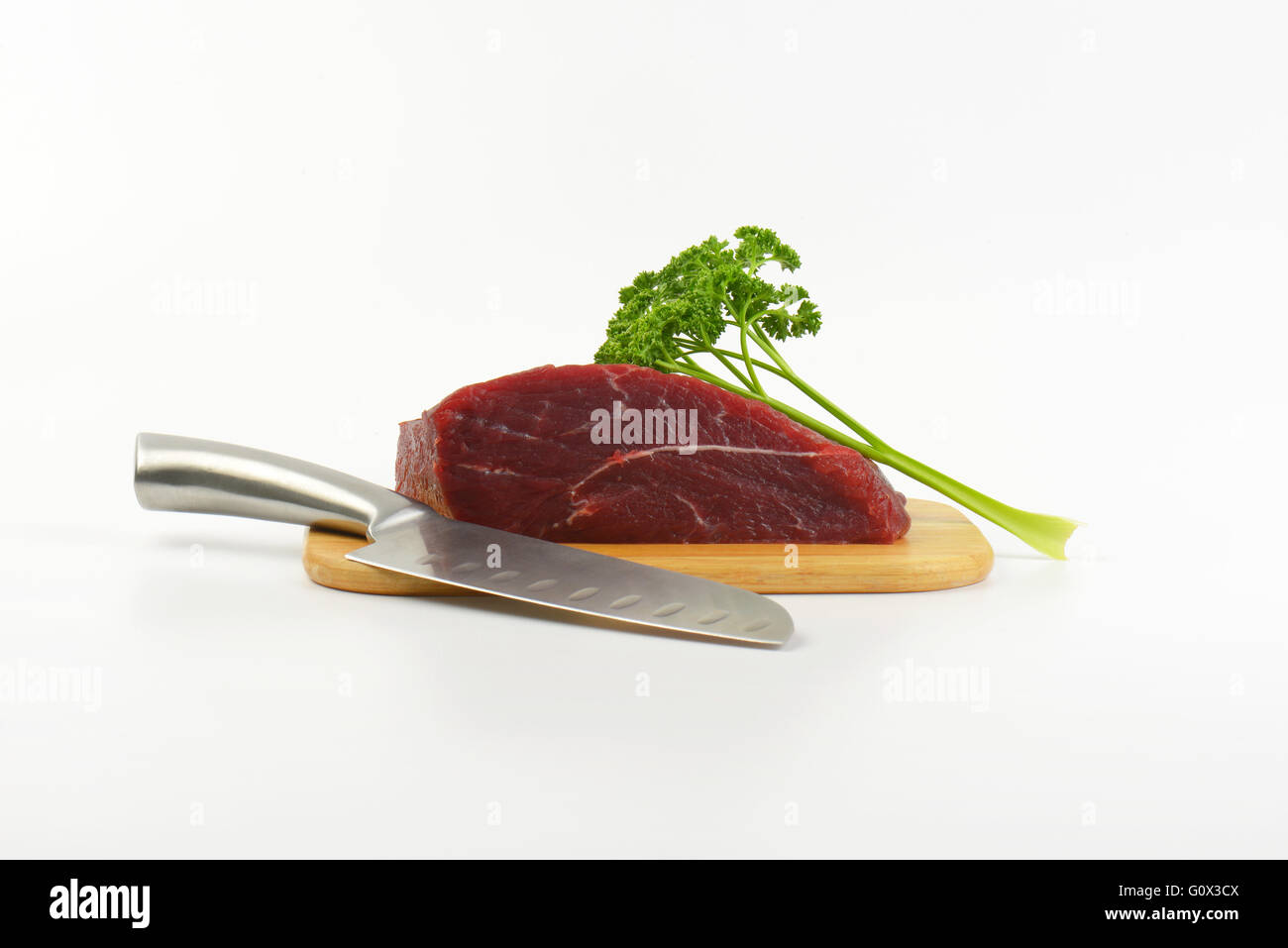 chunk of raw beef meat, parsley and sharp knife on wooden cutting board ...