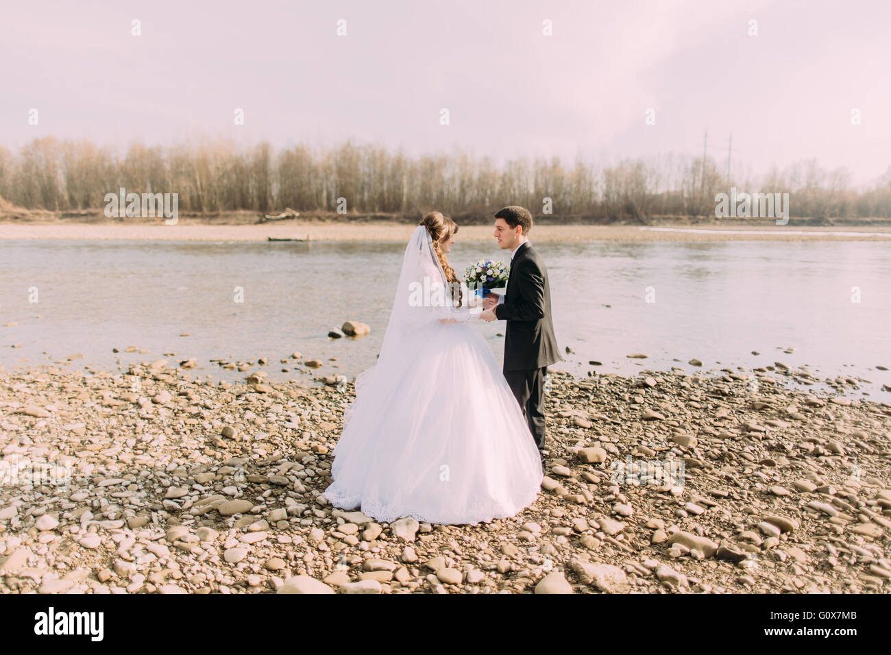Elegant gentle stylish groom and bride holding hands near river with ...