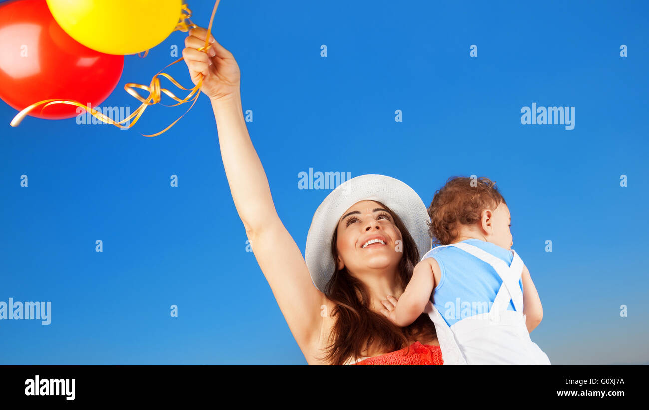 Portrait of a happy mother holding her cute little son in one hand and many colorful balloons in ...