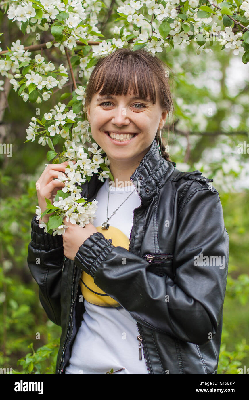 Beautiful smiling young woman in leather jacket standing near the blooming apple tree. Portrait ...