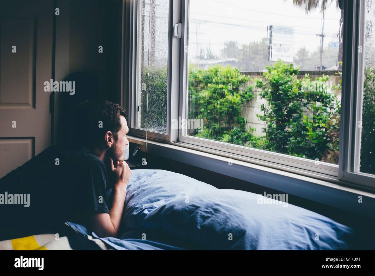 Man lying on bed staring through window at rain Stock Photo - Alamy