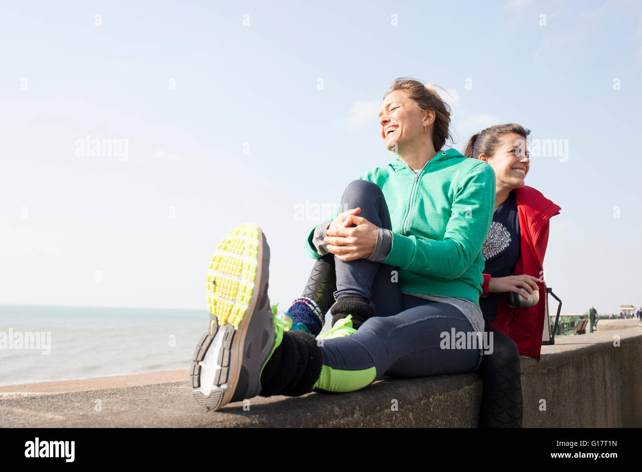 Two female runners taking a coffee break on wall at Brighton beach ...