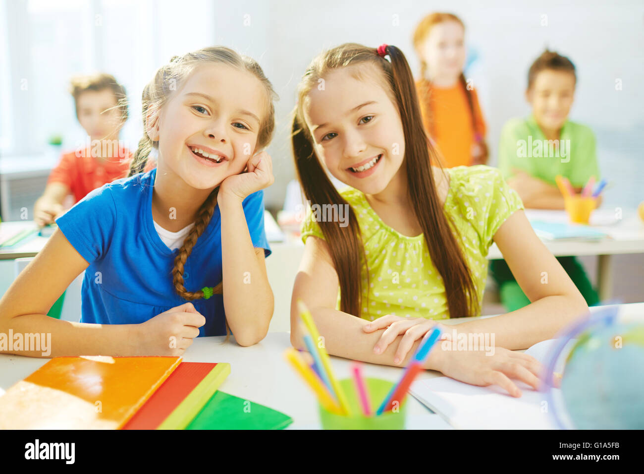 Friendly classmates sitting by desk and looking at camera with smiles ...