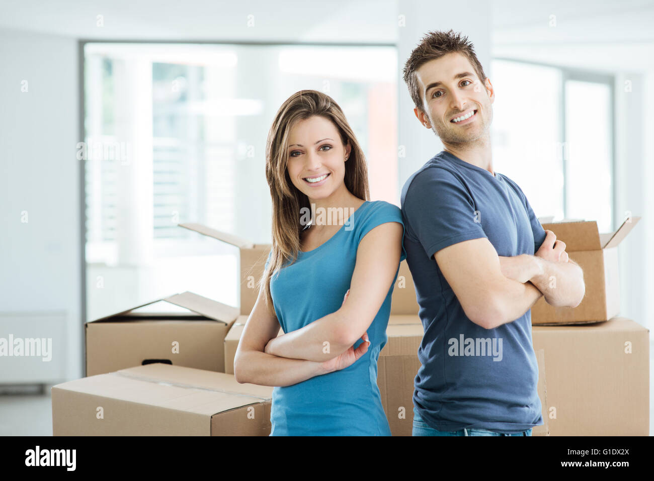 Smiling loving couple posing in their new house back to back surrounded ...