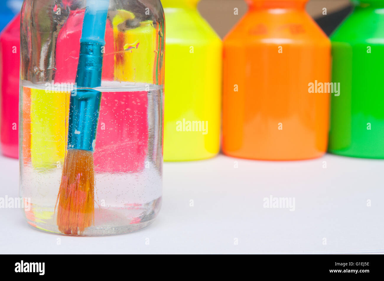 Paint brush in water in front of brightly coloured children's paint ...