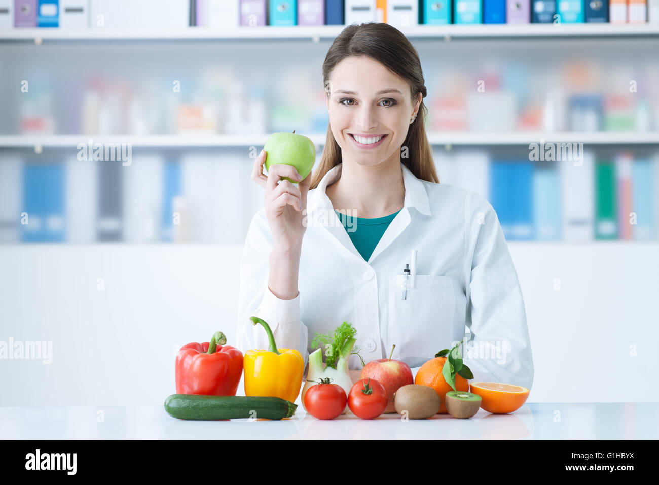 Smiling nutritionist in her office, she is holding a green apple and ...