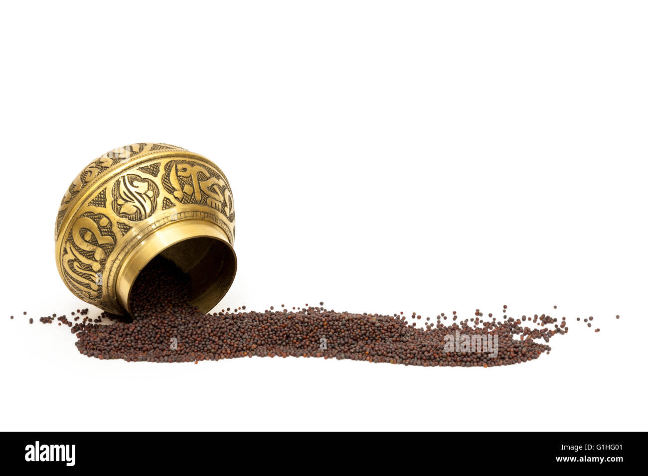 Whole brown mustard seeds tipped from an engraved metal spice bowl ...