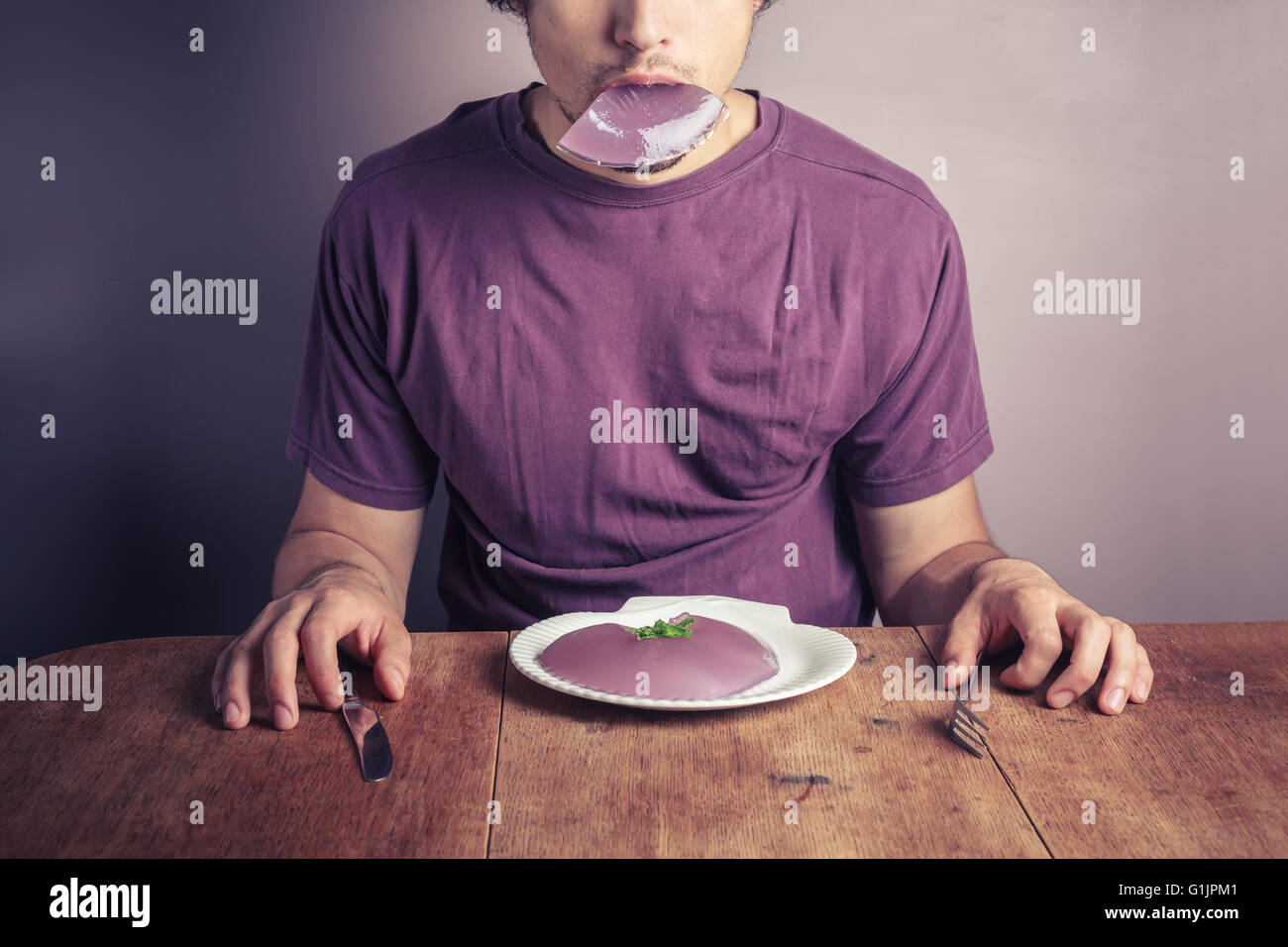 A young man is sitting at a table and eating a purple jelly pudding ...