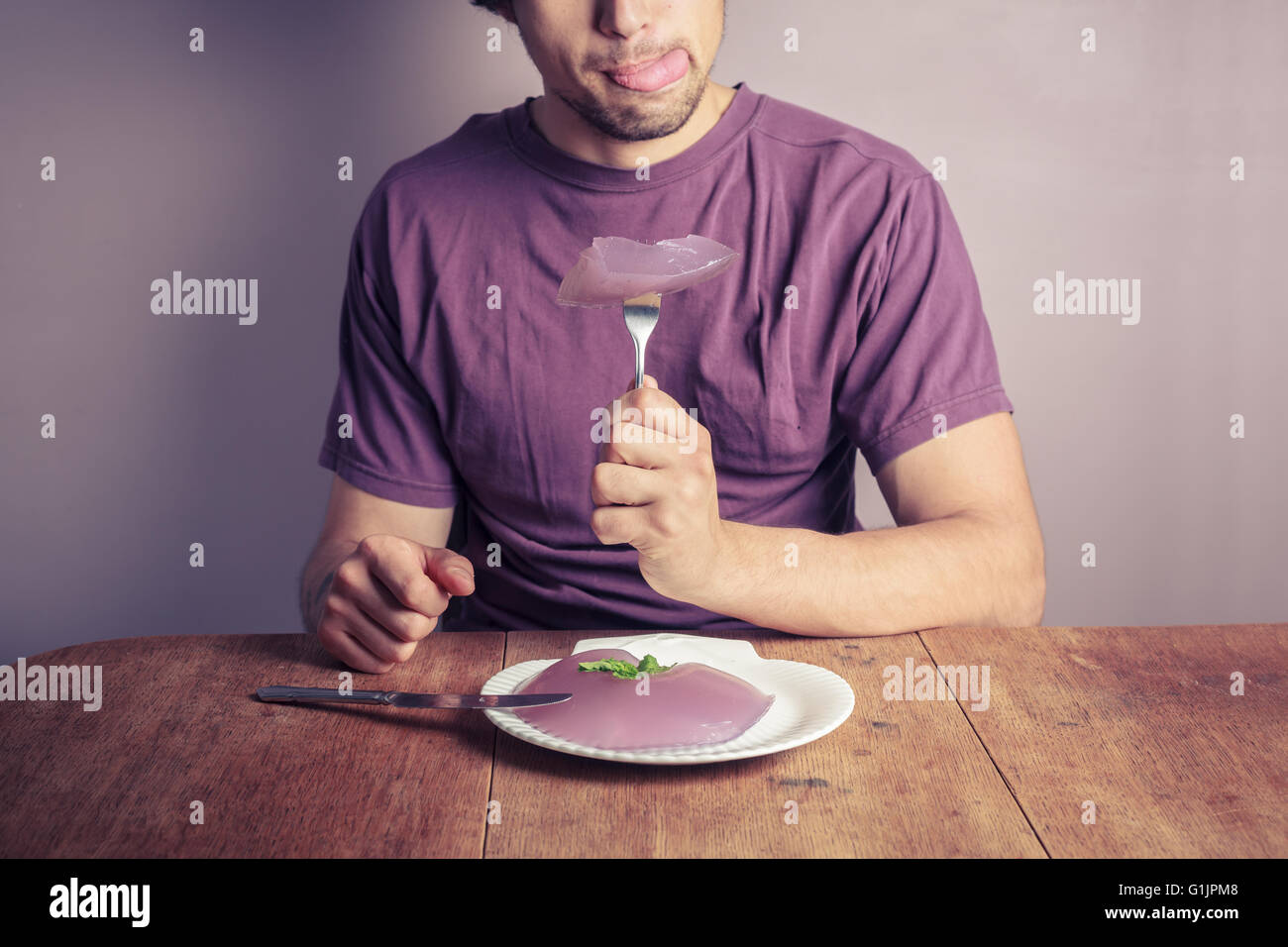 A young man is sitting at a table and eating a purple jelly pudding ...