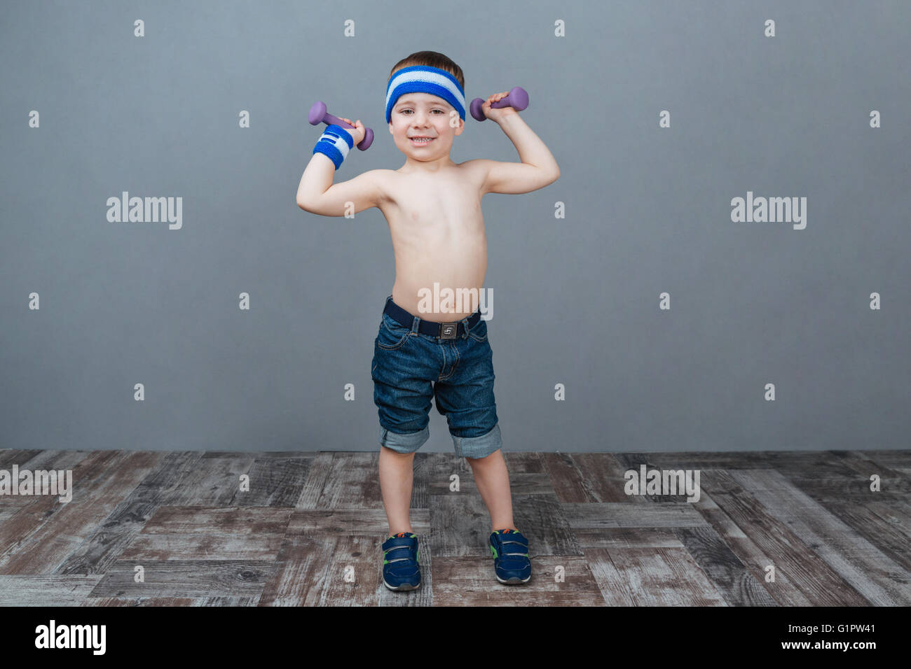 Smiling little boy doing exercises with dumbbells over grey background ...