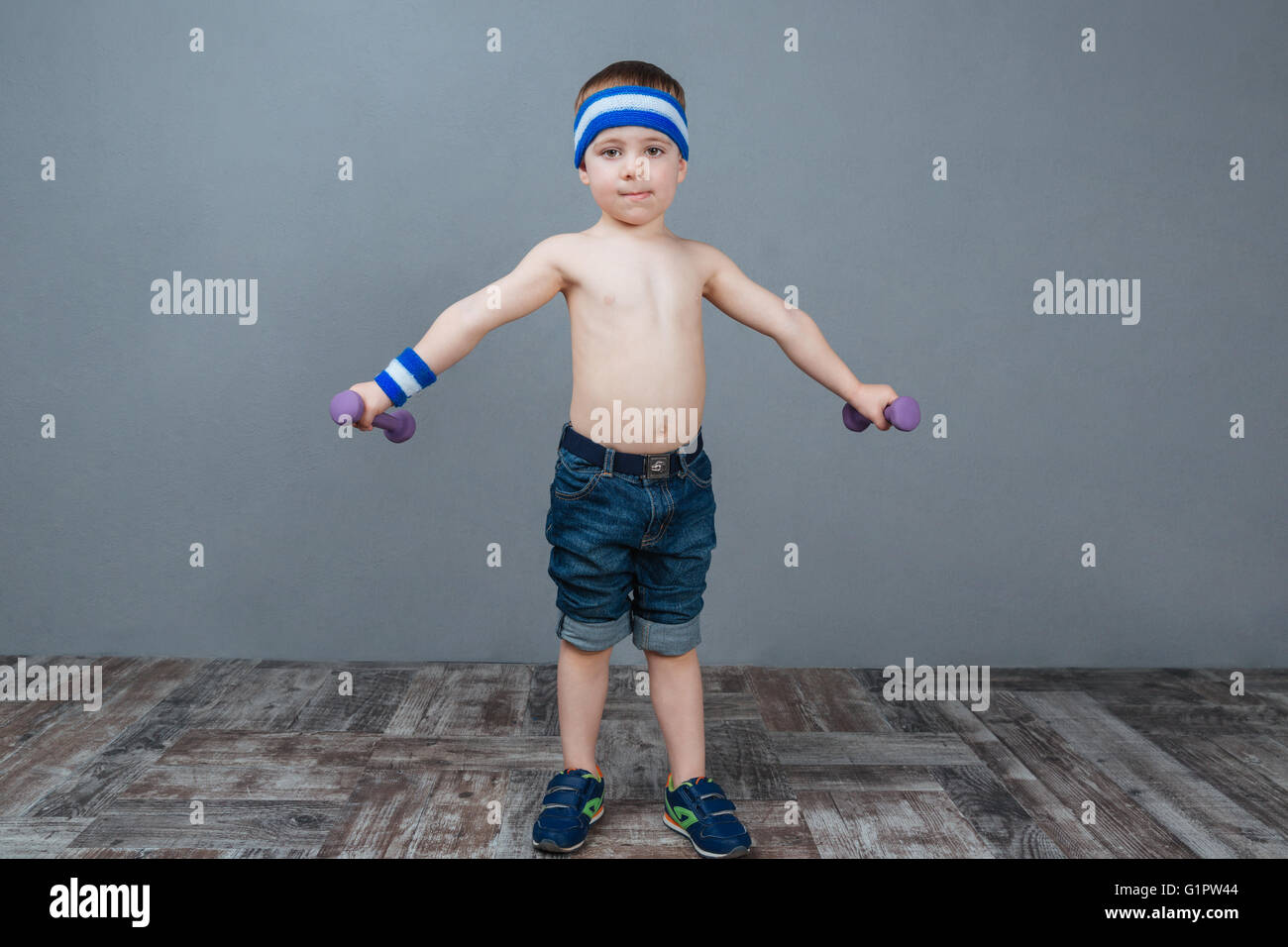 Cute little boy standing and working out using dumbbells over grey ...