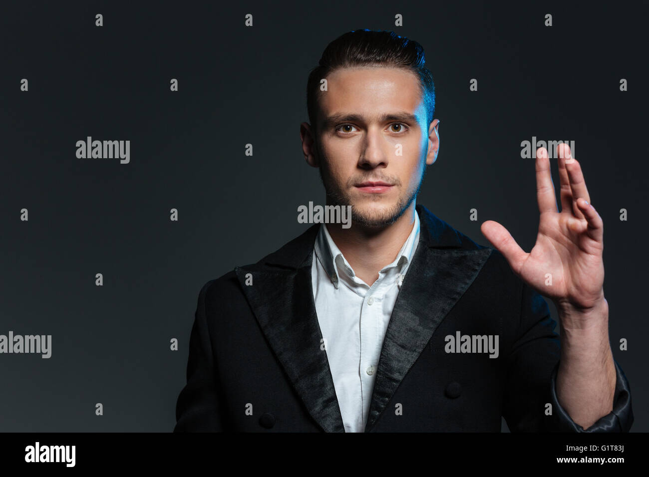 Portrait of confident young man magician with raised hand over grey ...