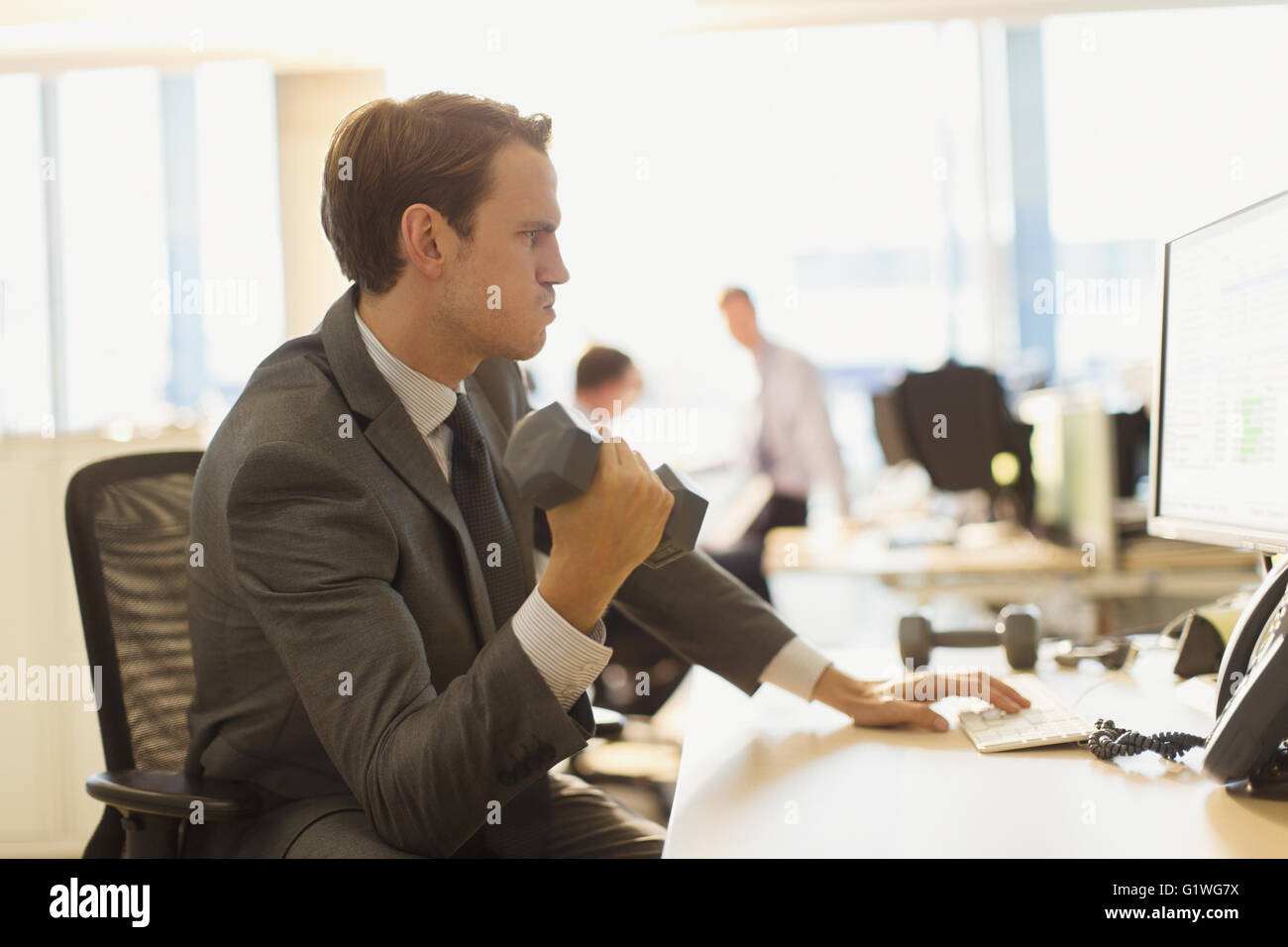 Businessman doing biceps curls with dumbbell at computer in office ...