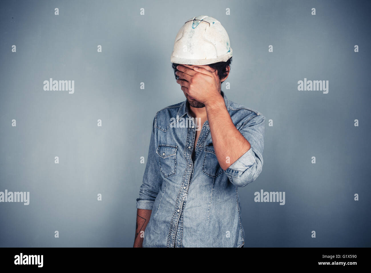 A blue collar worker wearing a hardhat is covering his face with his ...