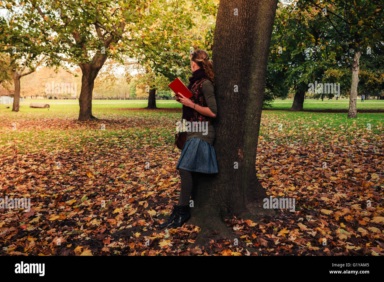 A young woman is leaning against a tree in the park and is reading a ...