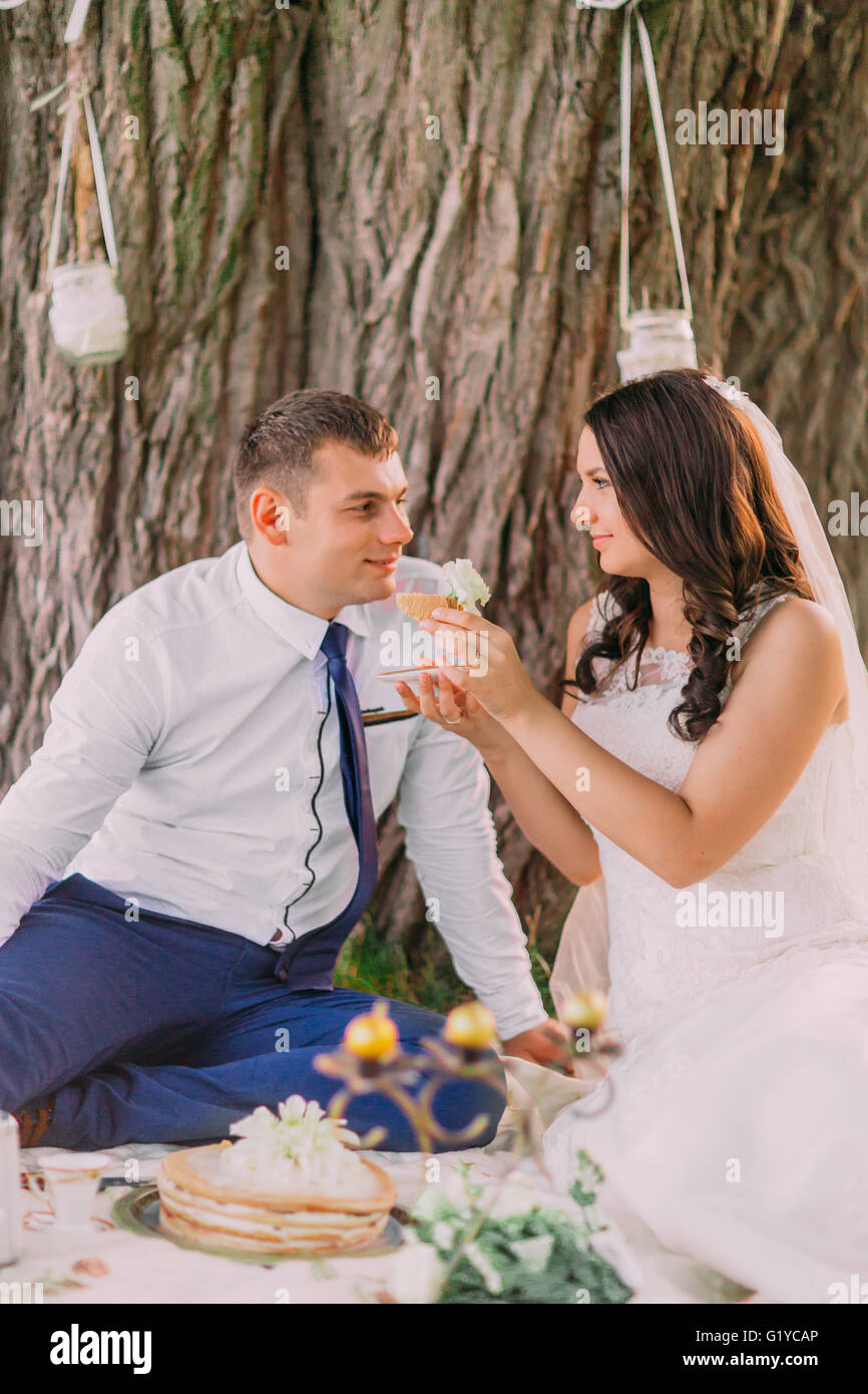 Beautiful young bride feeding wedding cake to groom outdoors on the ...