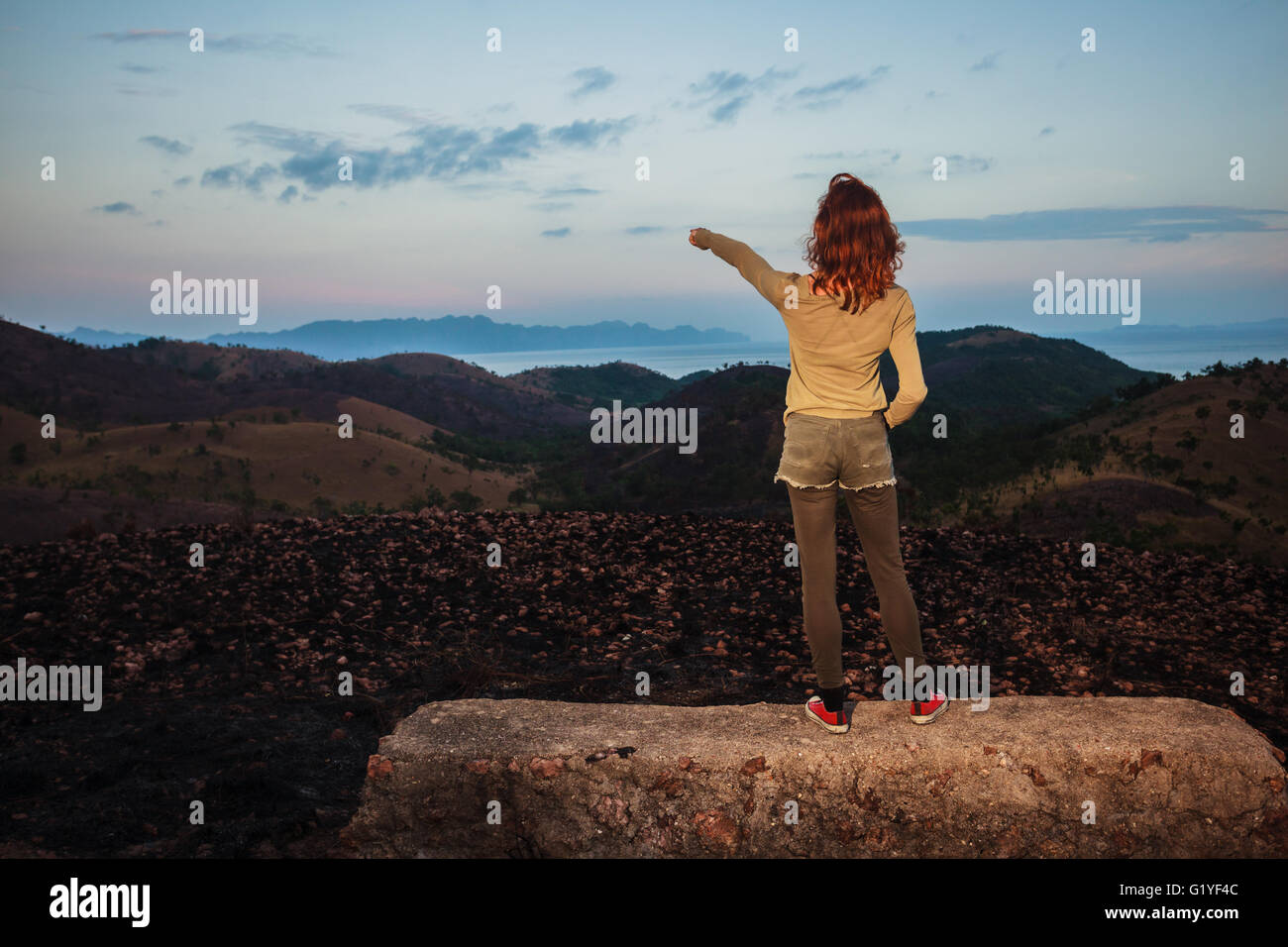 A young woman is standing on an unusual rock at sunset in a tropical ...