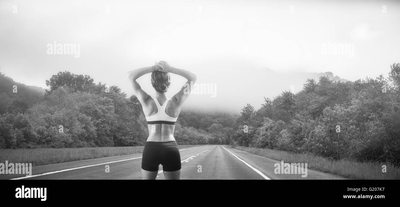 A Female runner stretching, looking down an empty stretch of road, shot ...