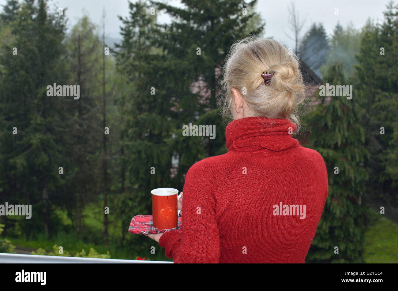 Woman relaxing, drinking tea or coffee and watching lush conifer trees ...