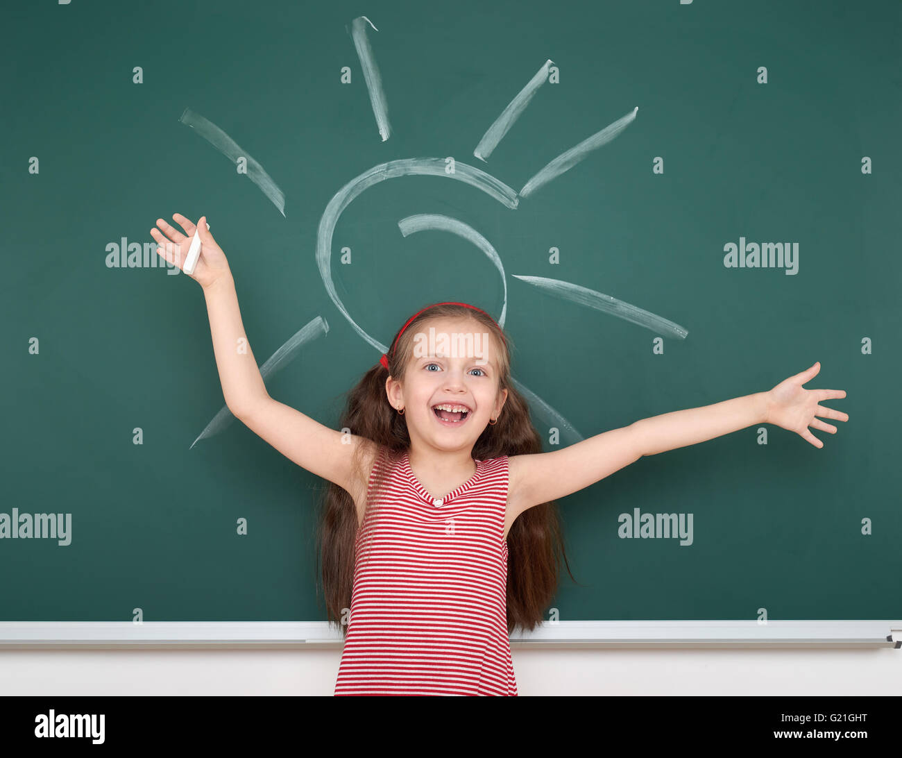 schoolgirl child in red striped dress drawing sun on green chalkboard ...