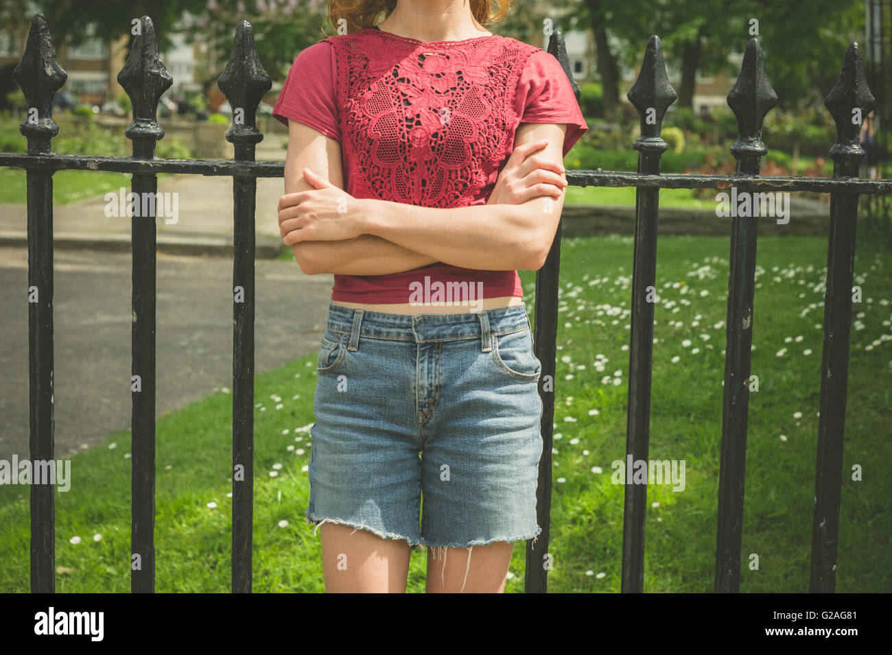 A young woman is standing by a gate outside a park in summer Stock ...