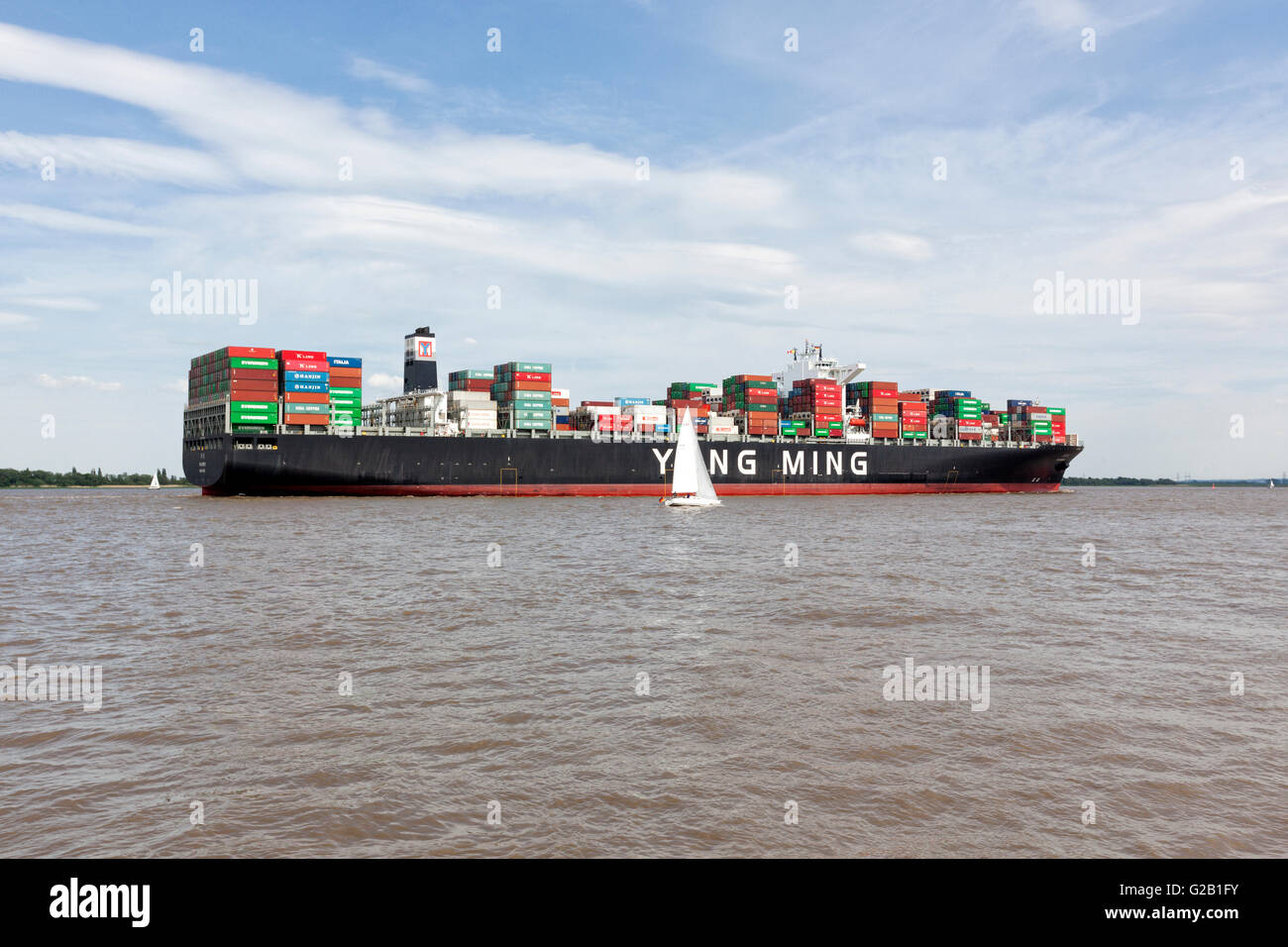 Ultra Large Container Ship YM Worth on the Elbe river near Hamburg. A small sail boat passing by ...