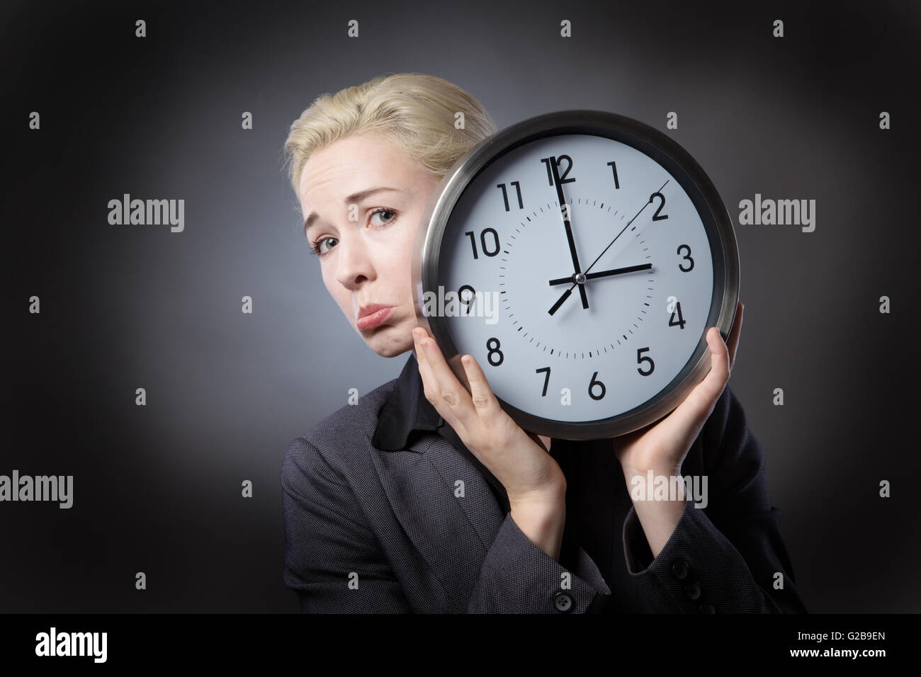 Woman in a suit with an unhappy expression, holding a large clock up by ...