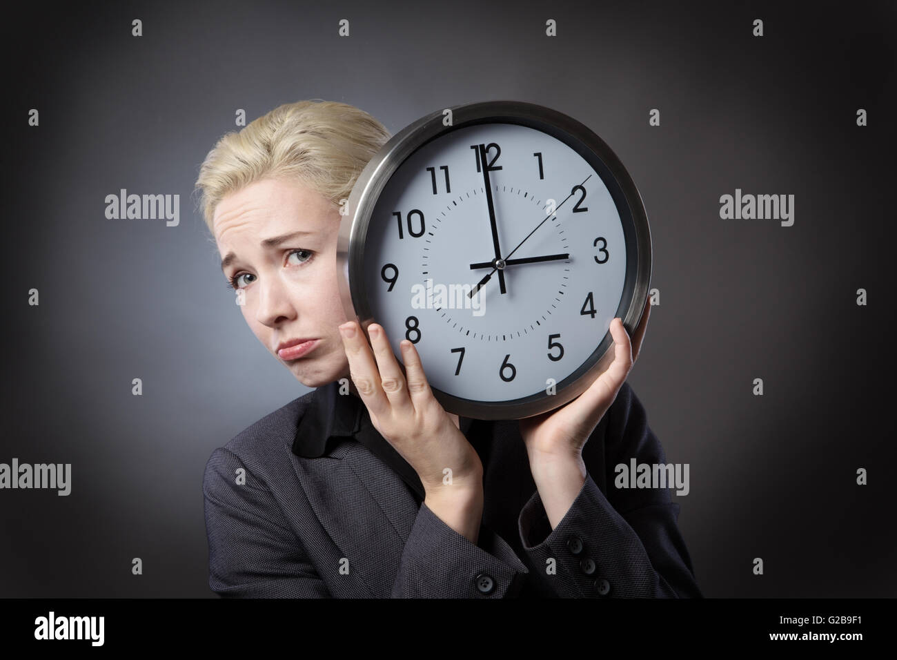 Woman in a suit with an unhappy expression, holding a large clock up by ...