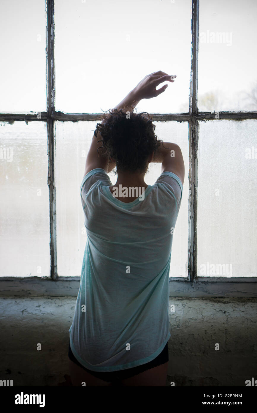 Rear view of a young woman standing by the window hands touching the ...