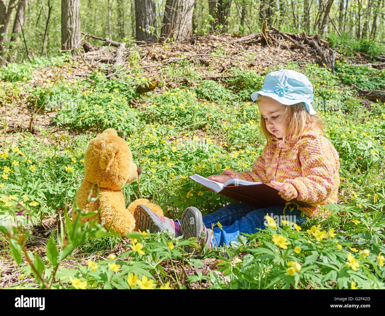 Cute Girl Reading Book Teddy Bear On The Grass In Forest Flower Glade