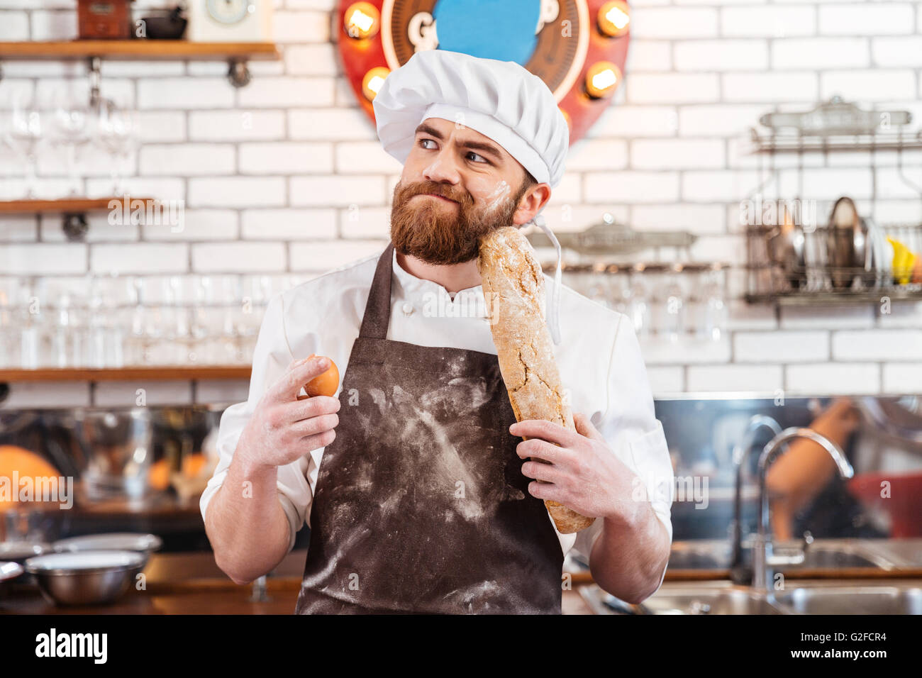 Pensive bearded baker holding eggs and bread and thinking on the ...